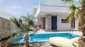 Uma área de piscina moderna com uma parede de pedra, palmeiras e uma espreguiçadeira bege.