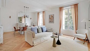 Uma sala de estar ampla, com sofá, cadeiras, espaço para refeições e grandes janelas com cortinas.