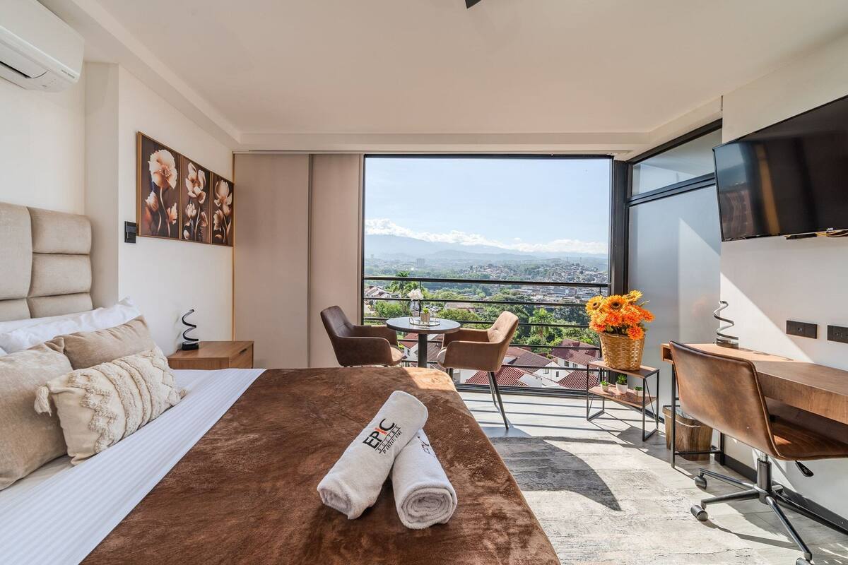 Quarto de hotel com uma cama, uma escrivaninha, uma cadeira, uma televisão e varanda com vista para a cidade e montanhas.