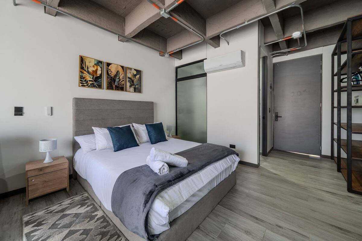 Quarto de hotel moderno com uma cama grande, mesa de cabeceira, ar condicionado fixado na parede e porta que dá para um corredor.
