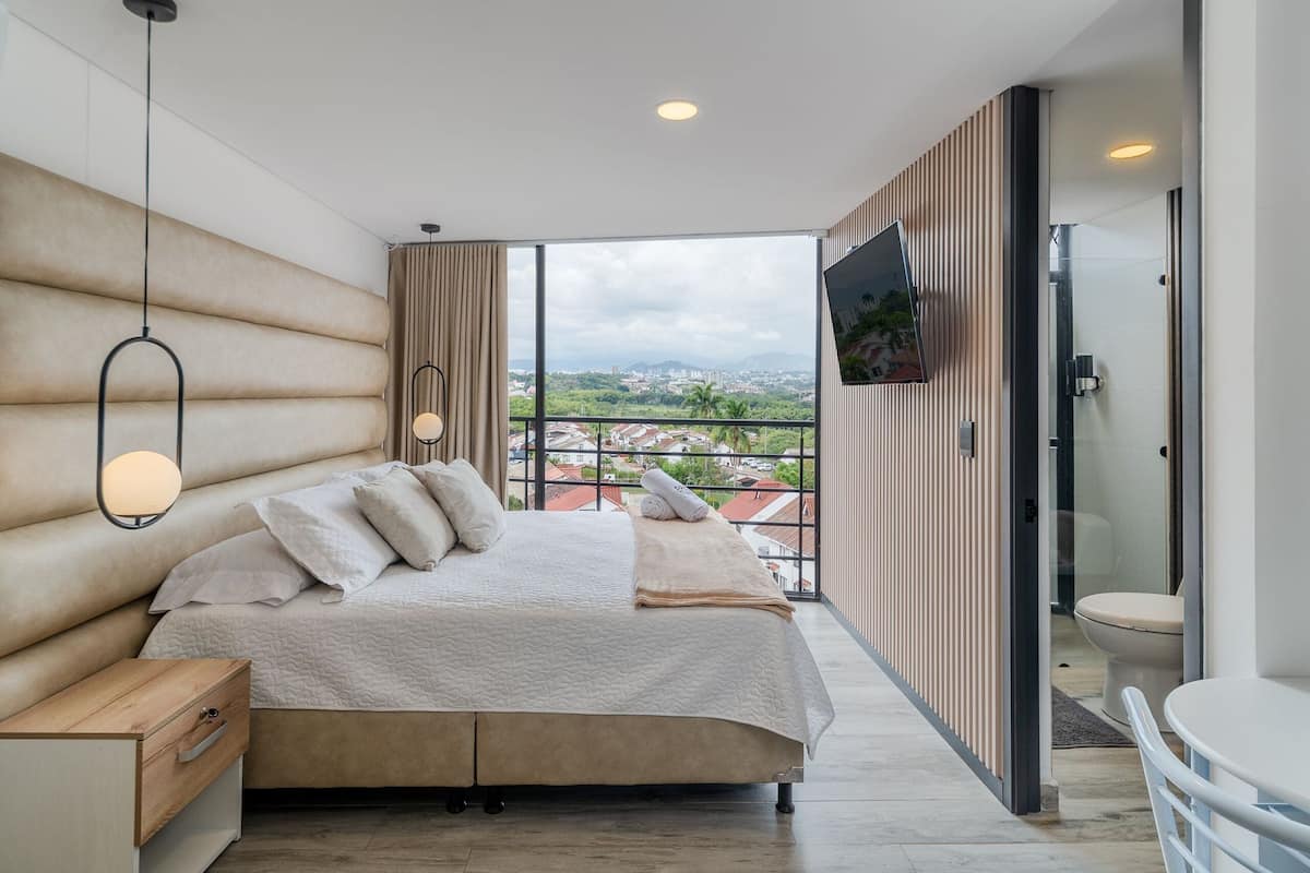 Quarto moderno com uma cama grande, mesas de cabeceira, televisão e varanda com vista para a cidade.