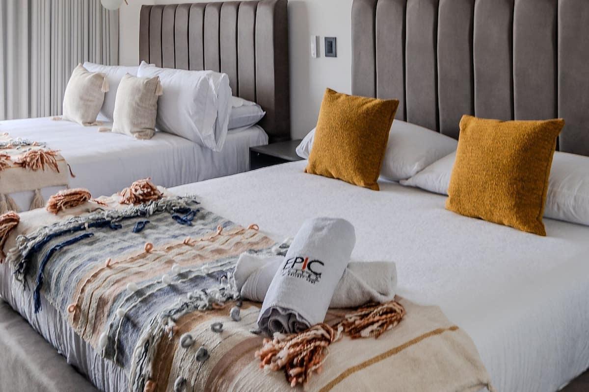 Quarto de hotel com duas camas, uma janela grande com cortinas e um abajur moderno.