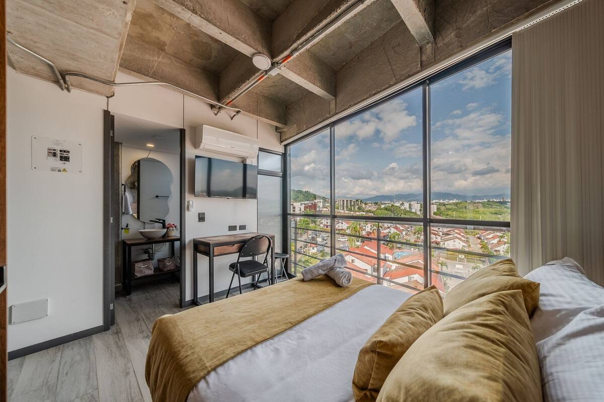 Quarto de hotel moderno com uma grande janela, uma cama, uma escrivaninha e um banheiro.