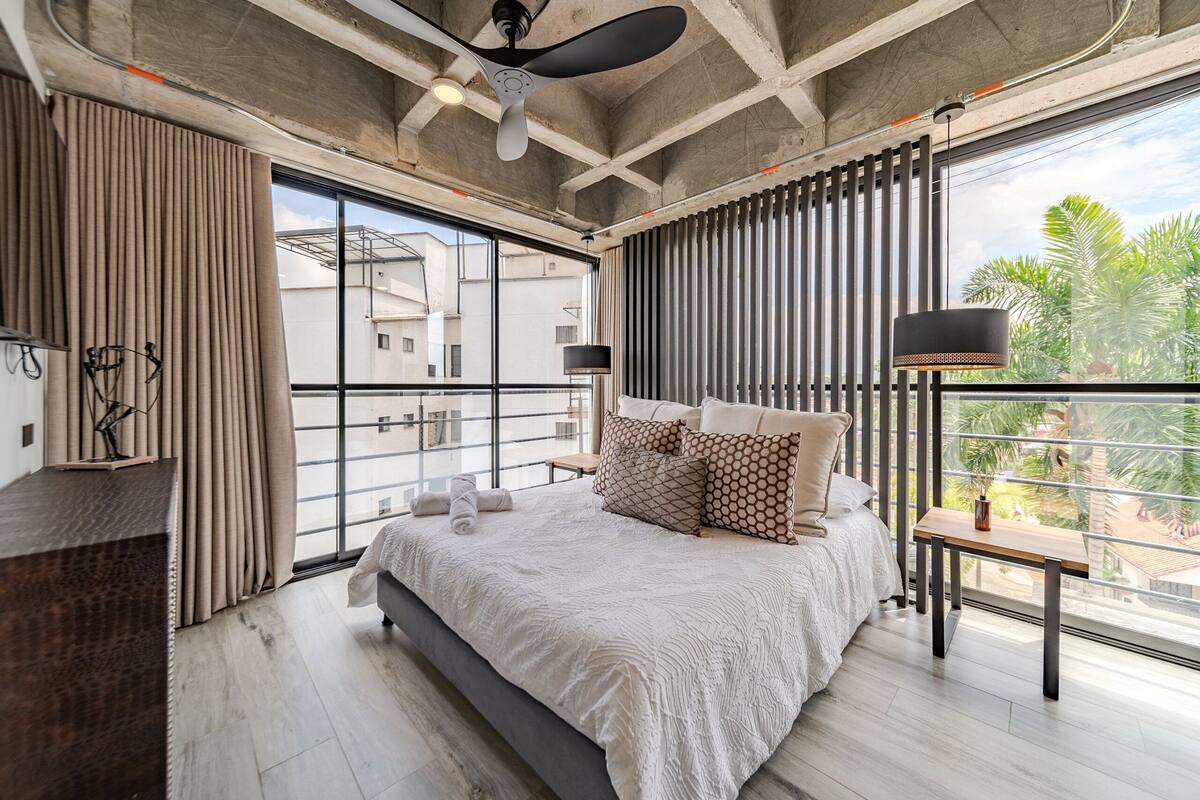Quarto moderno com uma cama grande, uma escrivaninha e uma varanda com vista para edifícios e palmeiras.