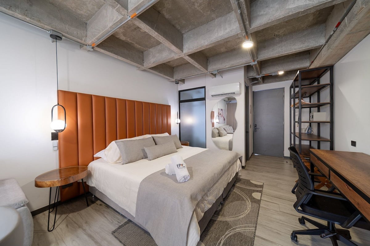 Quarto de hotel moderno com uma cama grande, cabeceira de madeira, escrivaninha e estante de livros.