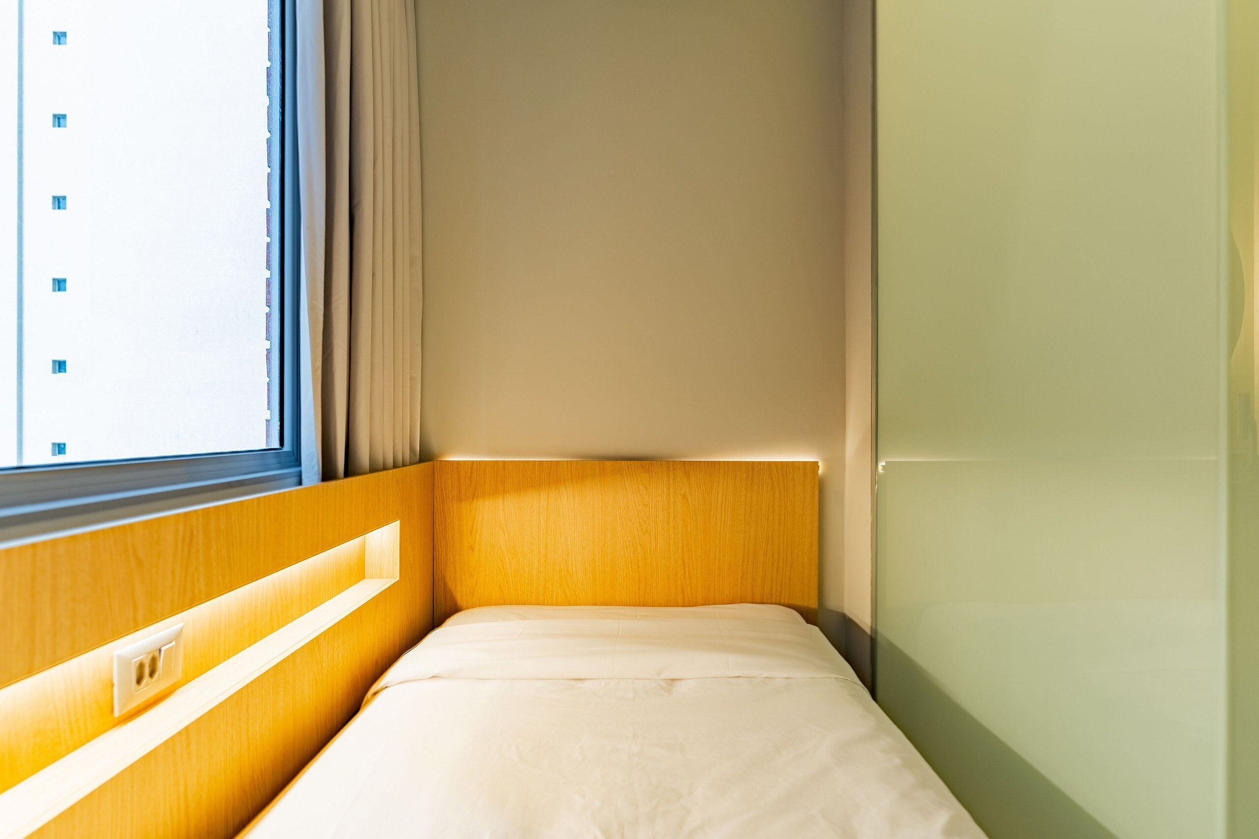 Quarto pequeno com uma cama de solteiro, cabeceira de madeira, janela com cortinas e parede verde.