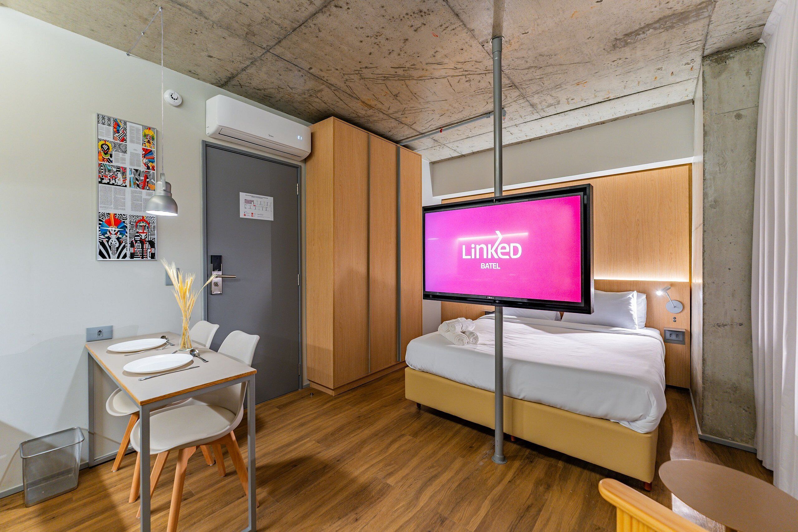 Quarto de hotel com cama, escrivaninha com cadeiras, televisão exibindo 'Linked' e parede com pôsteres.