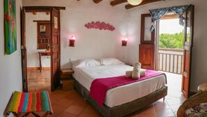 Quarto com cama, um banco de madeira, porta que dá para outro cômodo e varanda com vista.