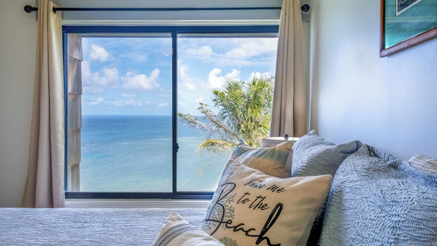 Quarto com uma grande janela com vista para o oceano, cama com várias almofadas e uma planta no parapeito da janela.