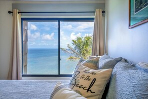 Quarto com uma grande janela com vista para o oceano, cama com várias almofadas e uma planta no parapeito da janela.