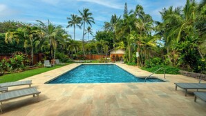 Piscina retangular com deck revestido, rodeada por palmeiras e vegetação exuberante.