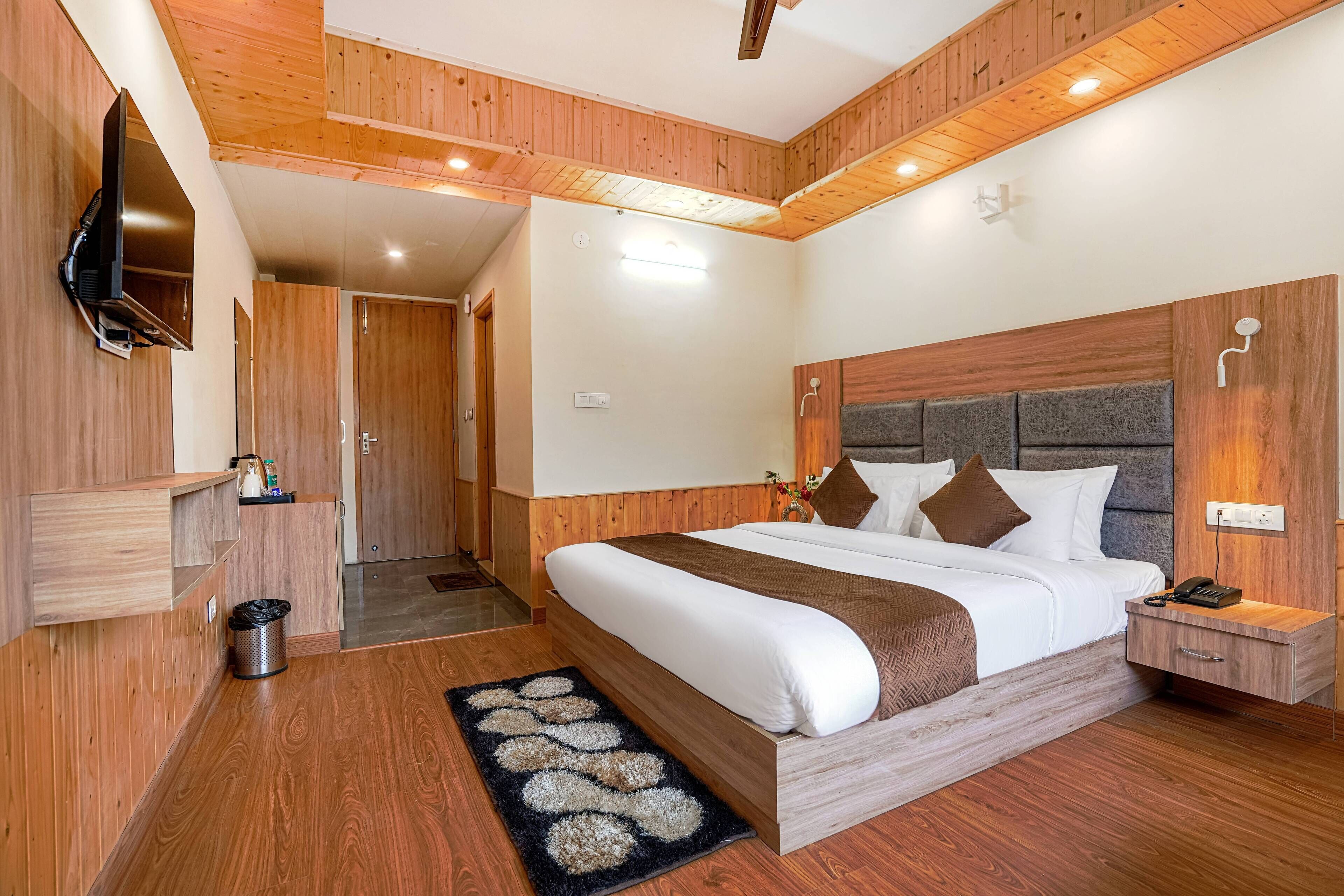 Quarto moderno com cabeceira de madeira, cama grande com roupa de cama branca, mesa de cabeceira, TV fixada na parede e teto de madeira com iluminação embutida.
