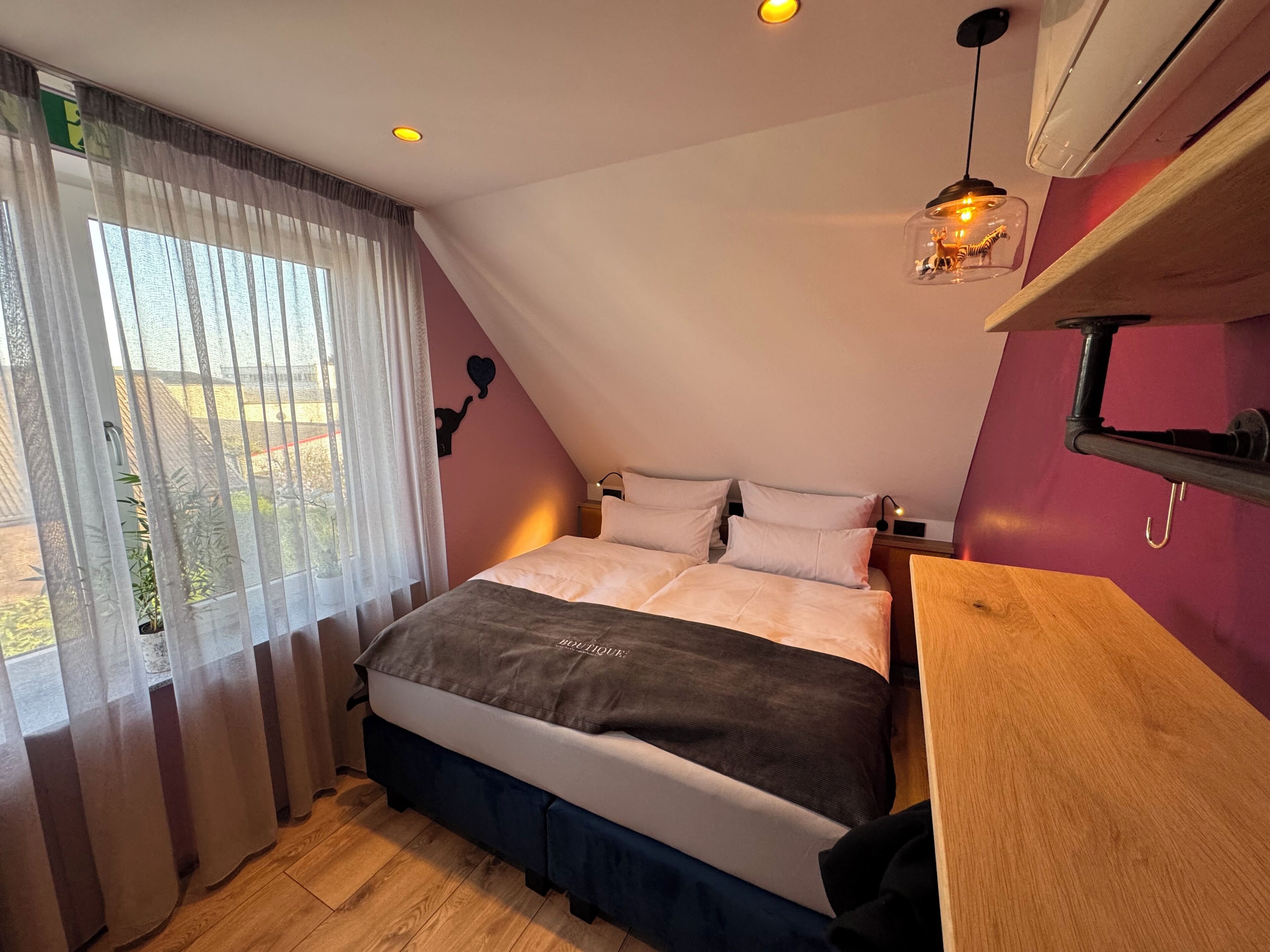 Quarto com cama, mesa de cabeceira de madeira, uma mesinha, janela com cortinas leves e ar-condicionado fixado na parede.