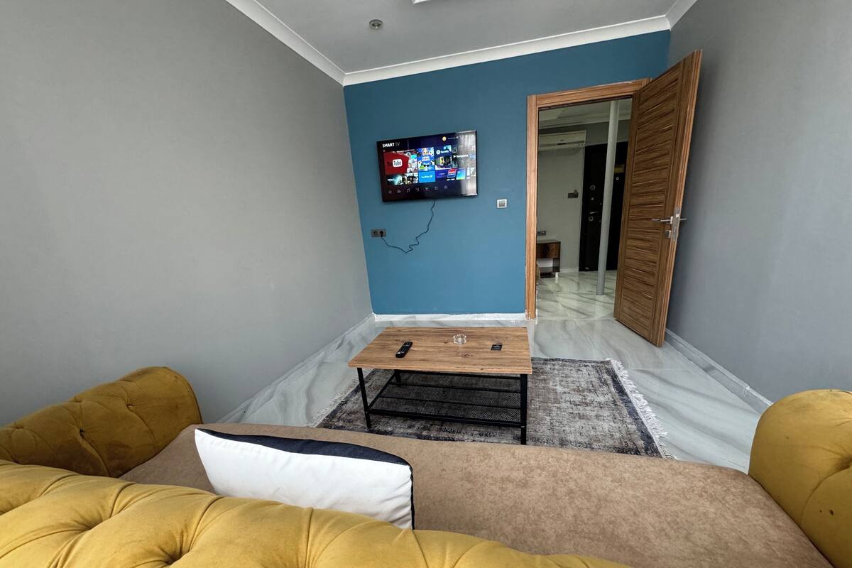 Quarto com parede azul decorativa, TV montada, mesa de madeira e sofá amarelo com detalhes embutidos.