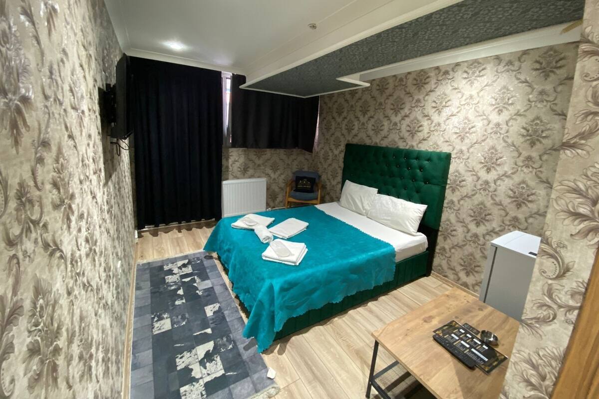 Quarto com cabeceira estofada verde, cama com roupa de cama branca, uma pequena mesa de madeira e um radiador.