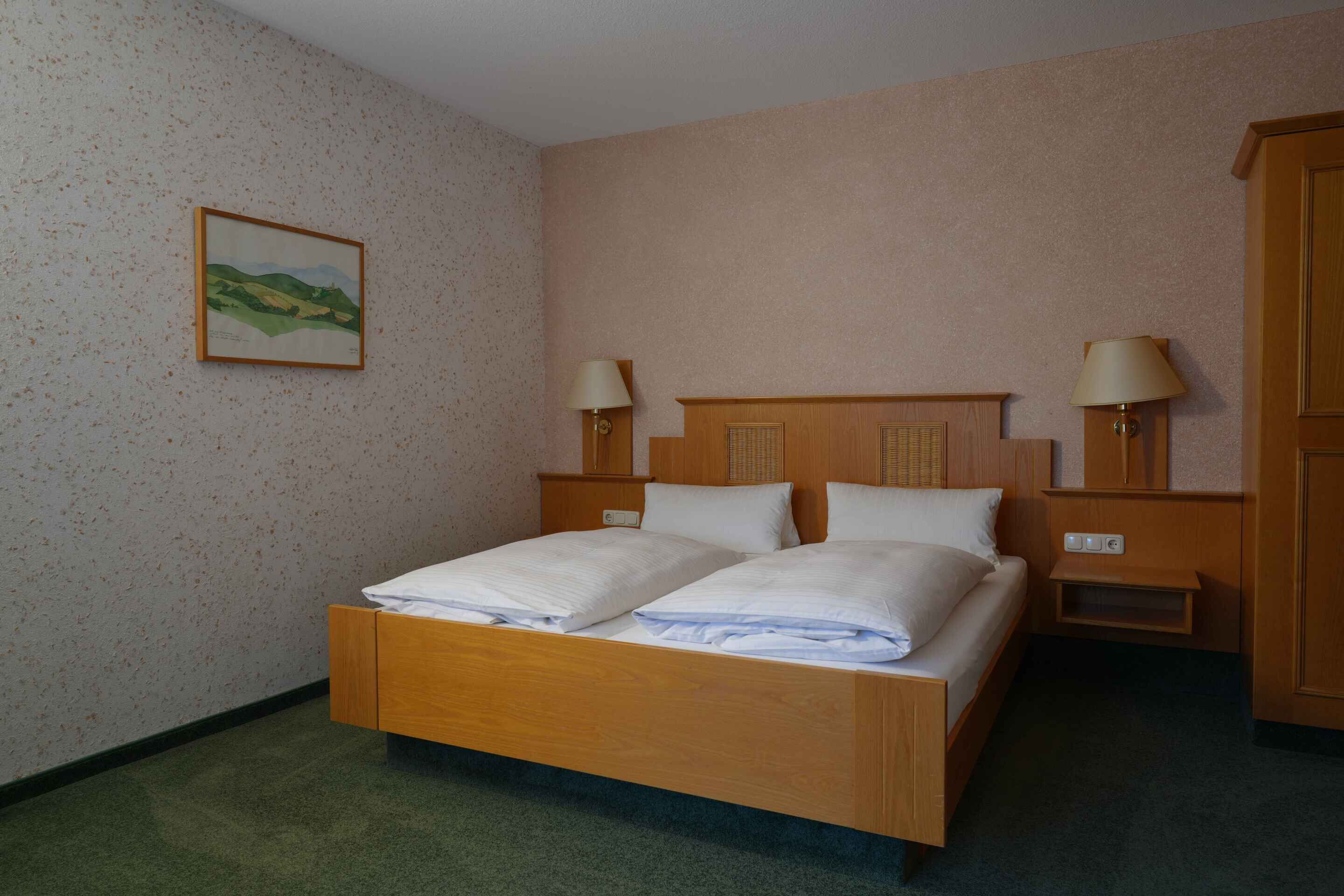 Quarto de hotel com cama de madeira, duas mesinhas de cabeceira com abajures, um quadro na parede e um tapete verde.