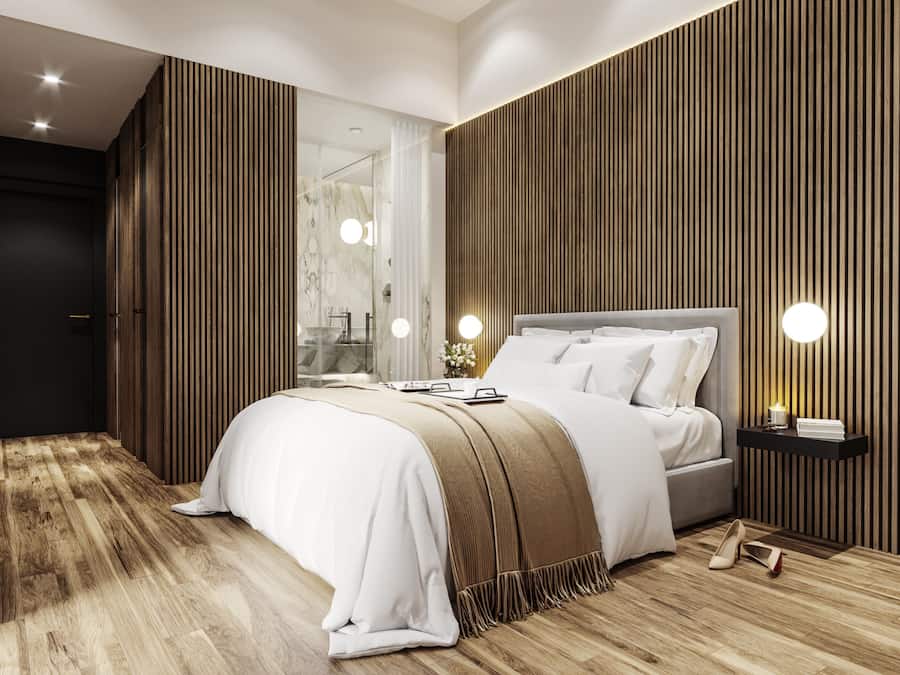 Quarto moderno com cama grande, painéis de madeira e banheiro de mármore visível através de uma porta de vidro.