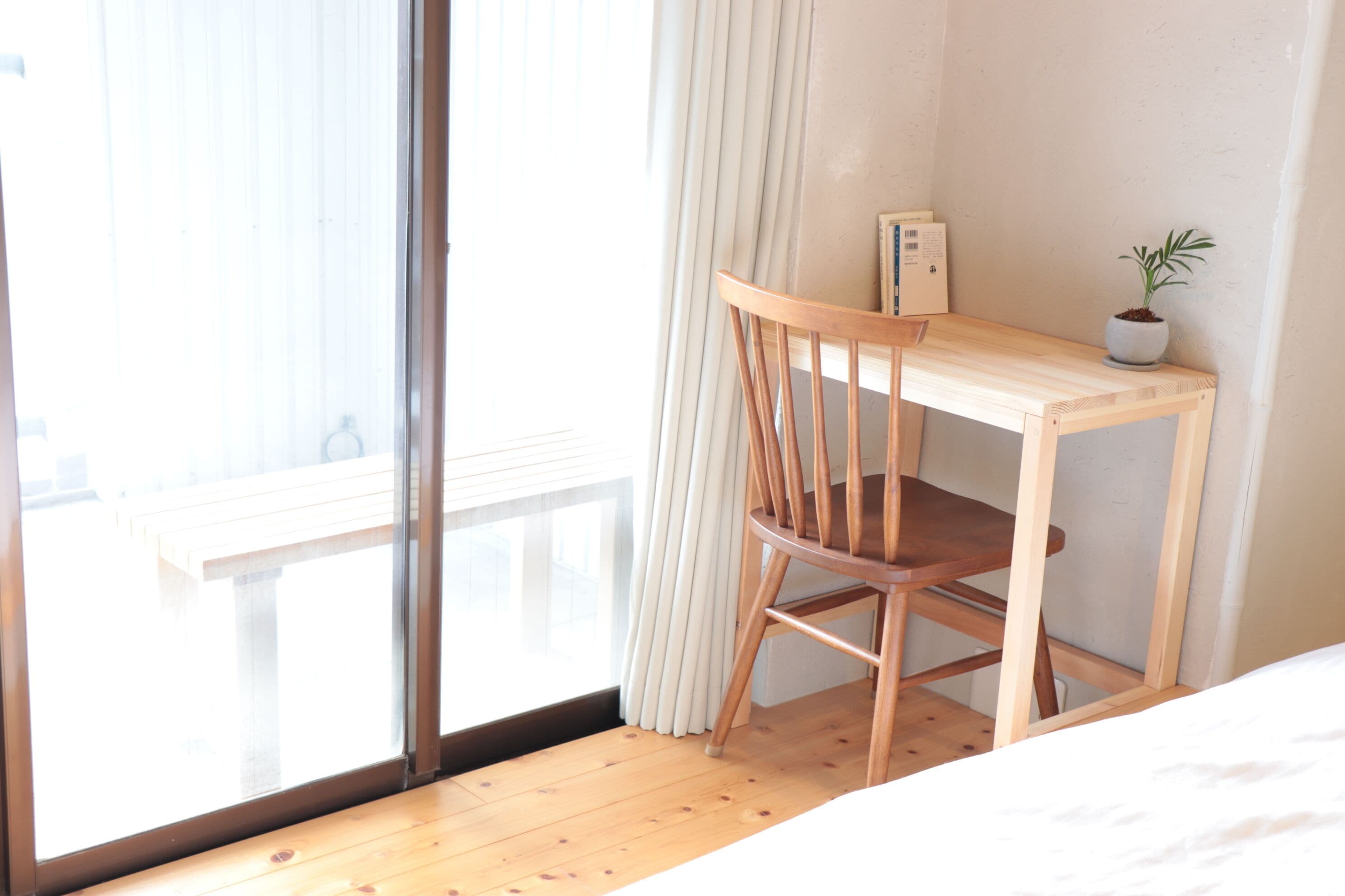 Quarto com escrivaninha de madeira, uma cadeira, planta em vaso e janela com cortinas.