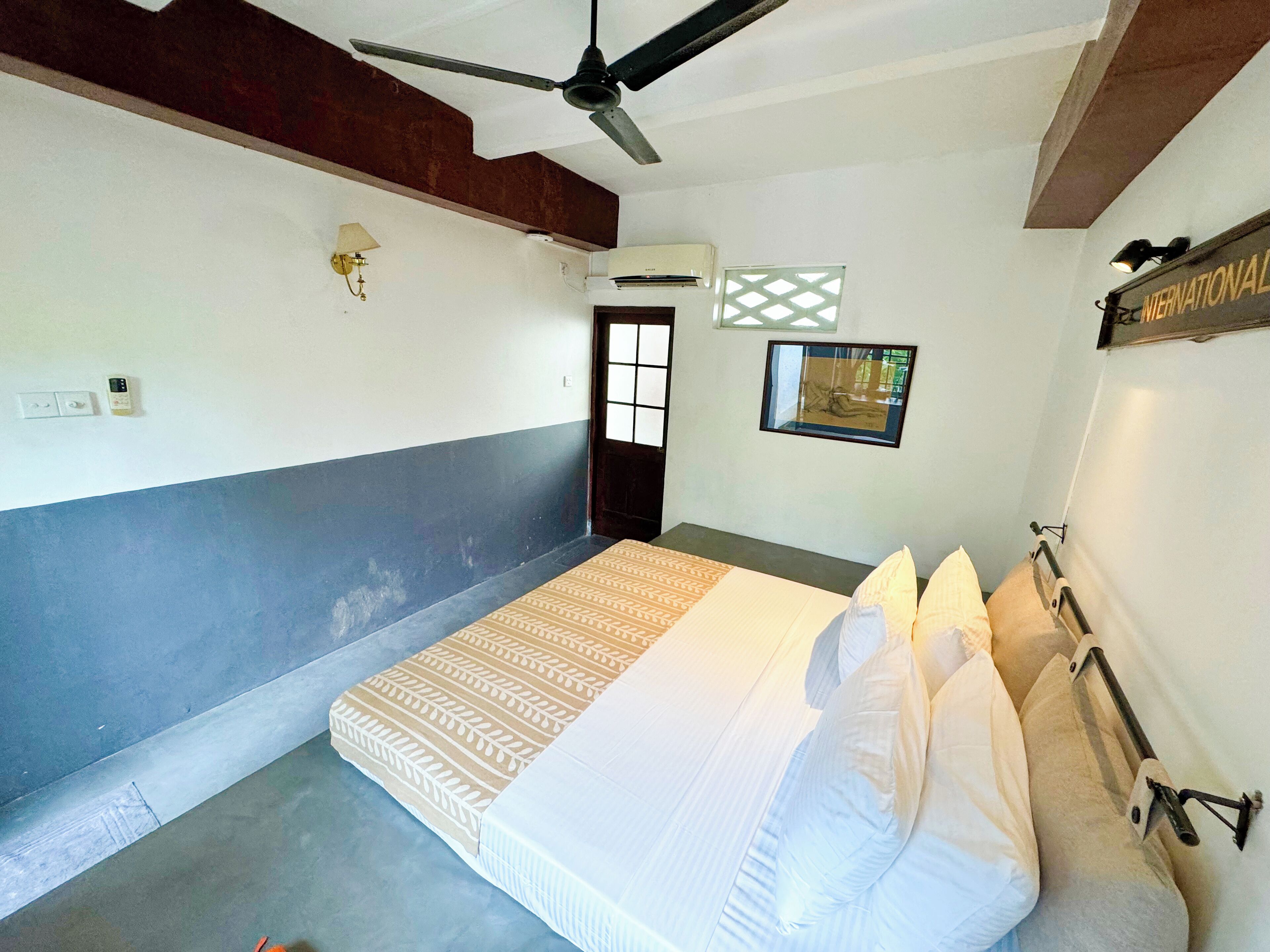 Quarto com cama, almofadas, ventoinha no teto, aparelho de ar condicionado, janela e luminária fixada na parede.