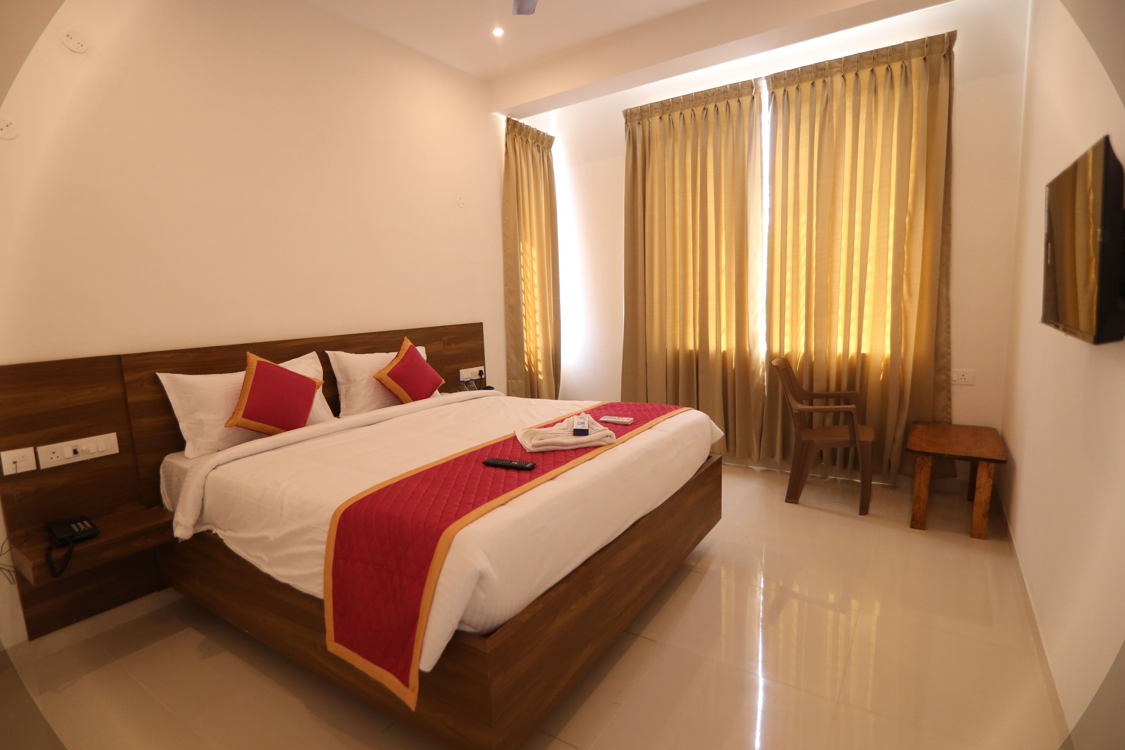 Quarto de hotel com cama de madeira, roupa de cama branca, detalhes em vermelho, mesa de cabeceira de madeira, uma cadeira e uma pequena mesa.
