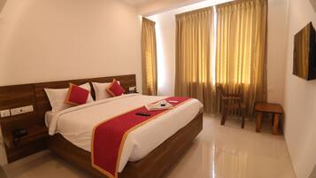 Quarto de hotel com cama de madeira, roupa de cama branca, detalhes em vermelho, mesa de cabeceira de madeira, uma cadeira e uma pequena mesa.