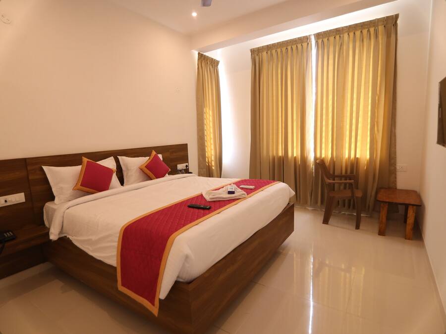 Quarto de hotel com cama de madeira, roupa de cama branca, detalhes em vermelho, mesa de cabeceira de madeira, uma cadeira e uma pequena mesa.