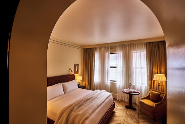Quarto de hotel com uma cama grande, uma mesinha com luminária, uma cadeira e uma janela com cortinas.