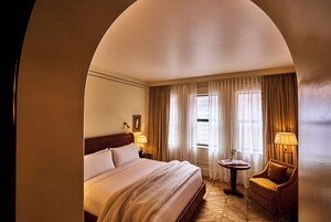 Quarto de hotel com uma cama grande, uma mesinha com luminária, uma cadeira e uma janela com cortinas.