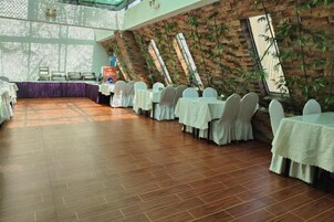 Uma sala de banquetes com mesas dispostas em fileiras, parede de tijolos com decoração em bambu e uma grande janela que permite a entrada de luz natural.