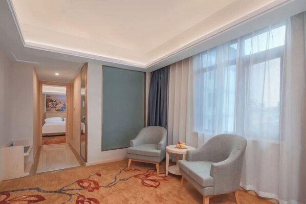 Quarto de hotel com uma cama, duas poltronas, uma mesinha e uma janela grande com cortinas.