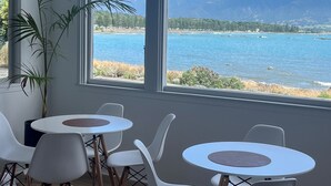 Quarto com grandes janelas que oferecem vista para um lago e montanhas. Há cadeiras e mesas brancas dispostas para refeições.