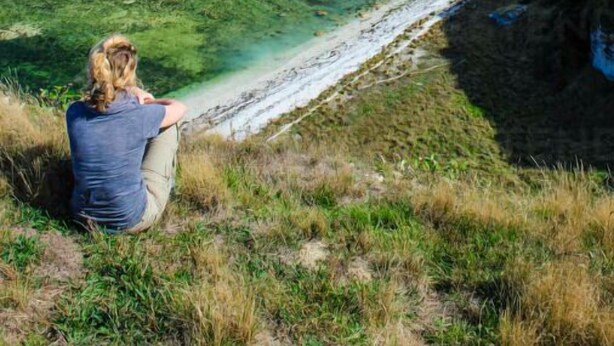 Uma pessoa sentada em uma colina gramada, com vista para uma paisagem costeira com águas claras e formações rochosas.