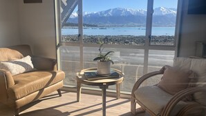 Quarto com uma janela ampla que oferece vista para um lago e montanhas. Há duas cadeiras, uma com almofada e outra com almofada e um franjário. Uma pequena mesa com uma planta e um livro está no centro.