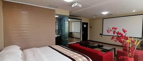 Quarto de hotel com uma cama, um sofá vermelho, uma mesinha e um projetor.