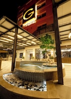 Um hotel com um logotipo 'G' em destaque, área de piscina com paisagismo, pedras e uma cascata, além de área de estar coberta ao ar livre.