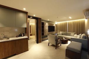 Uma sala de estar moderna com sofá, banquinho e mesa de centro. Há uma área de cozinha com pia e armários. O quarto tem uma janela grande com cortinas.