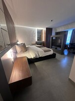 Quarto moderno com uma cama grande, um banco de madeira, uma escrivaninha e uma televisão.