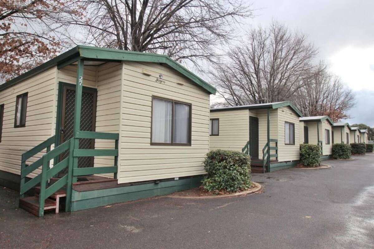 Uma fileira de pequenas cabanas térreas, com detalhes em verde e portas numeradas, dispostas em uma área pavimentada, com árvores ao fundo.