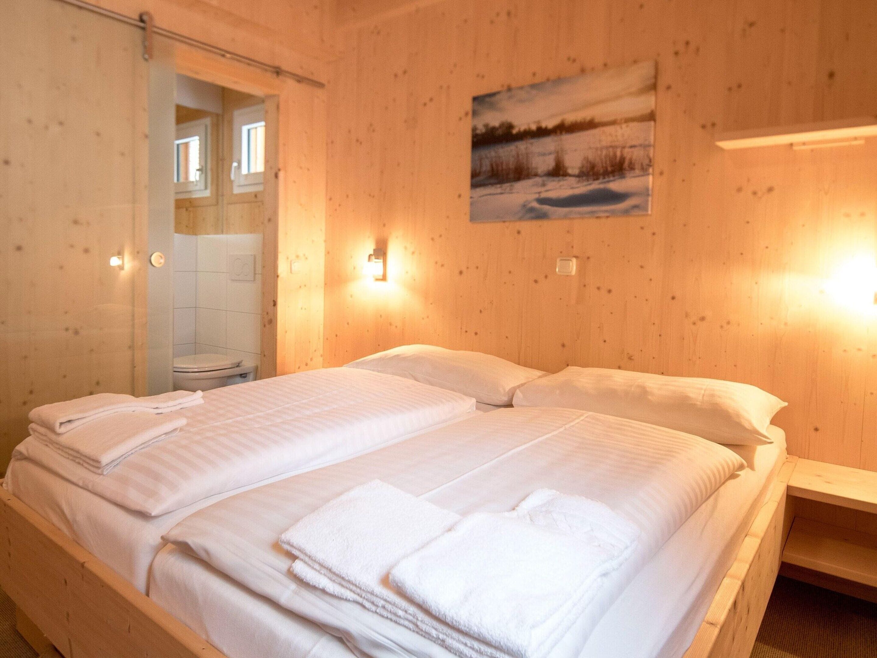Quarto de madeira com uma cama, um quadro e um banheiro visível através de uma porta de vidro.