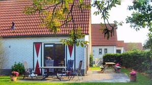 Uma casa com telhado em telhas, um pátio com cadeiras e uma mesa, e um jardim.