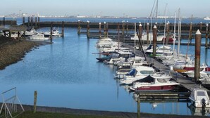 Uma marina com barcos atracados na beira da água, um calçadão de madeira e um horizonte urbano distante.