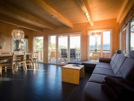 Interior de uma cabana rústica moderna, com área de jantar, sofá e grandes portas de correr que dão para um deck externo.