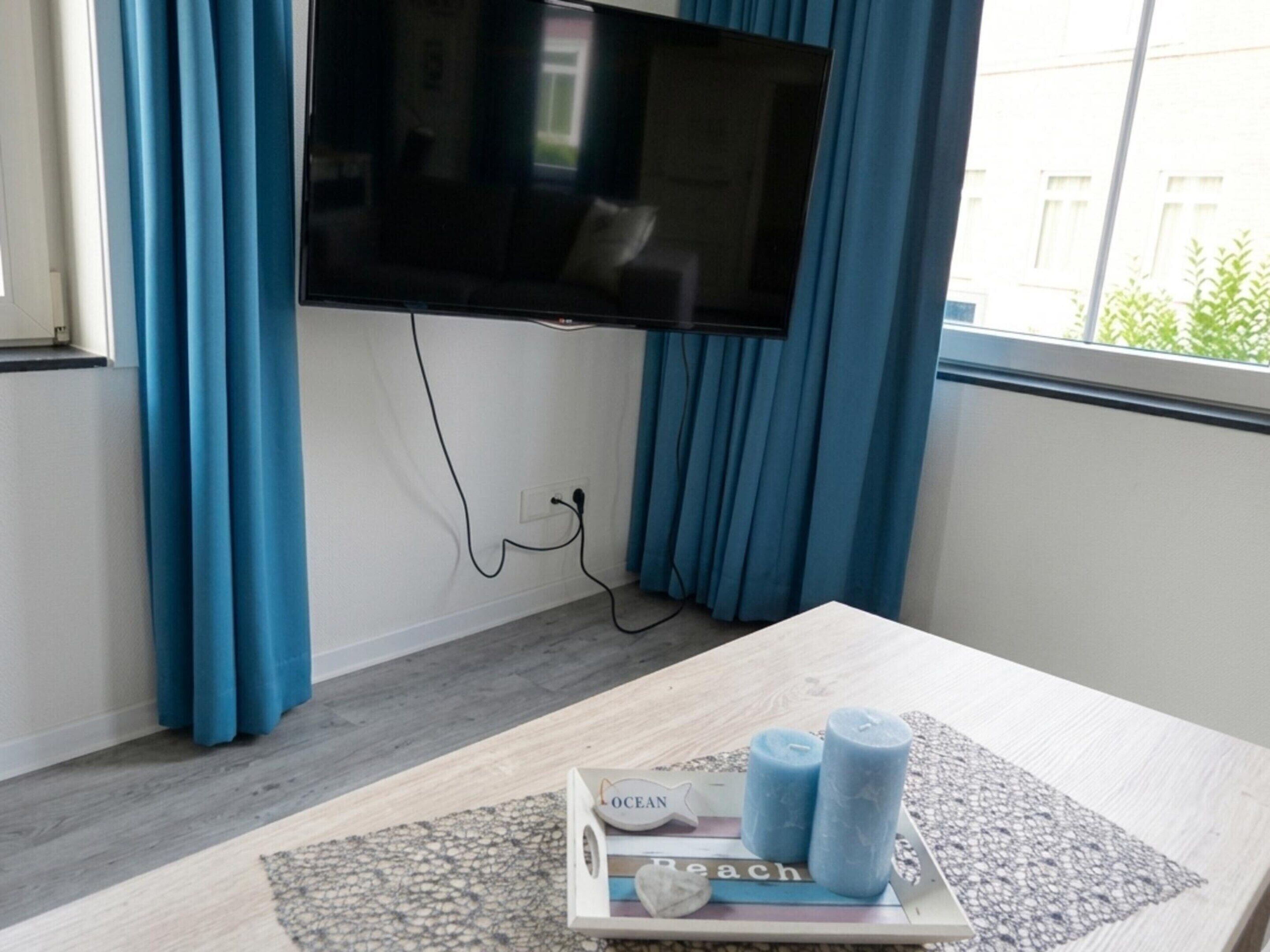 Quarto com televisão de tela plana na parede, mesa de madeira com toalha de mesa e janela com cortinas azuis.