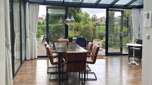 Uma área de refeições com mesa e cadeiras de vidro, uma garrafa sobre a mesa e vista para o verde lá fora.