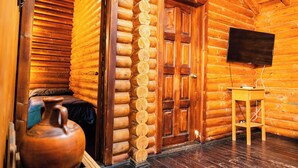 Interior de uma cabana de tronco com uma porta de madeira, uma televisão fixada na parede e uma pequena mesa de madeira.