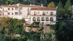 Um edifício branco com telhado ladrilhado, várias varandas e janelas em arco, rodeado por vegetação exuberante e terreno rochoso.