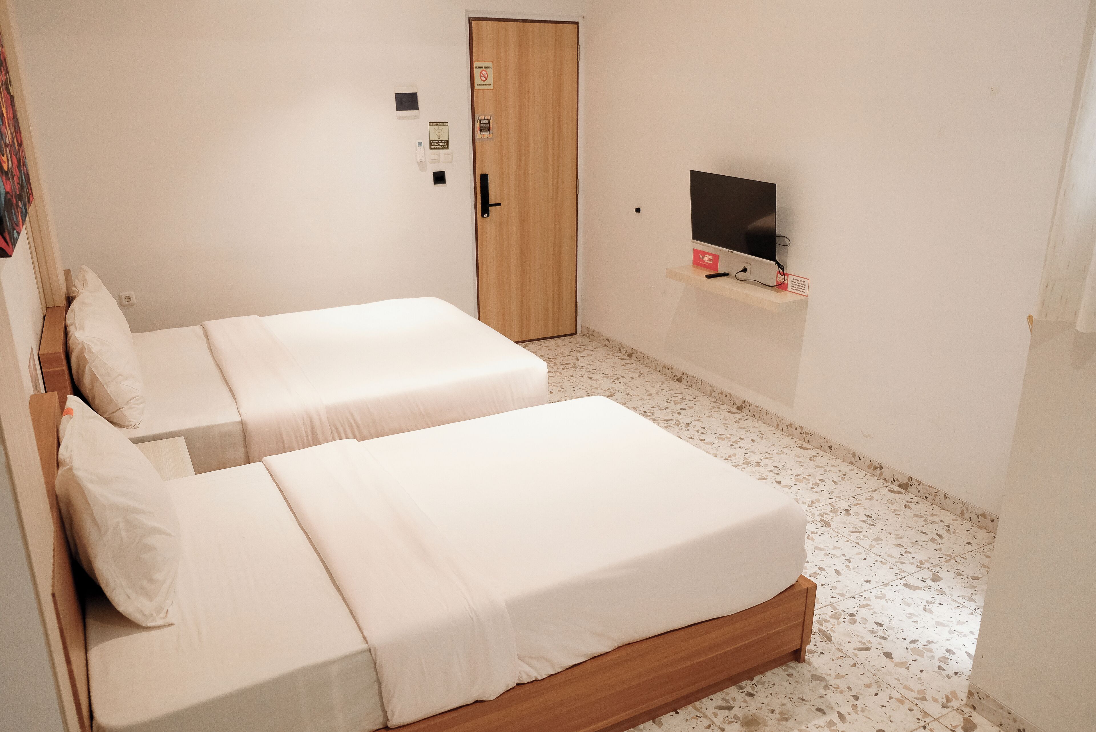 Quarto com duas camas de solteiro, porta de madeira, televisão em móvel e prateleira pequena com uma caixa.