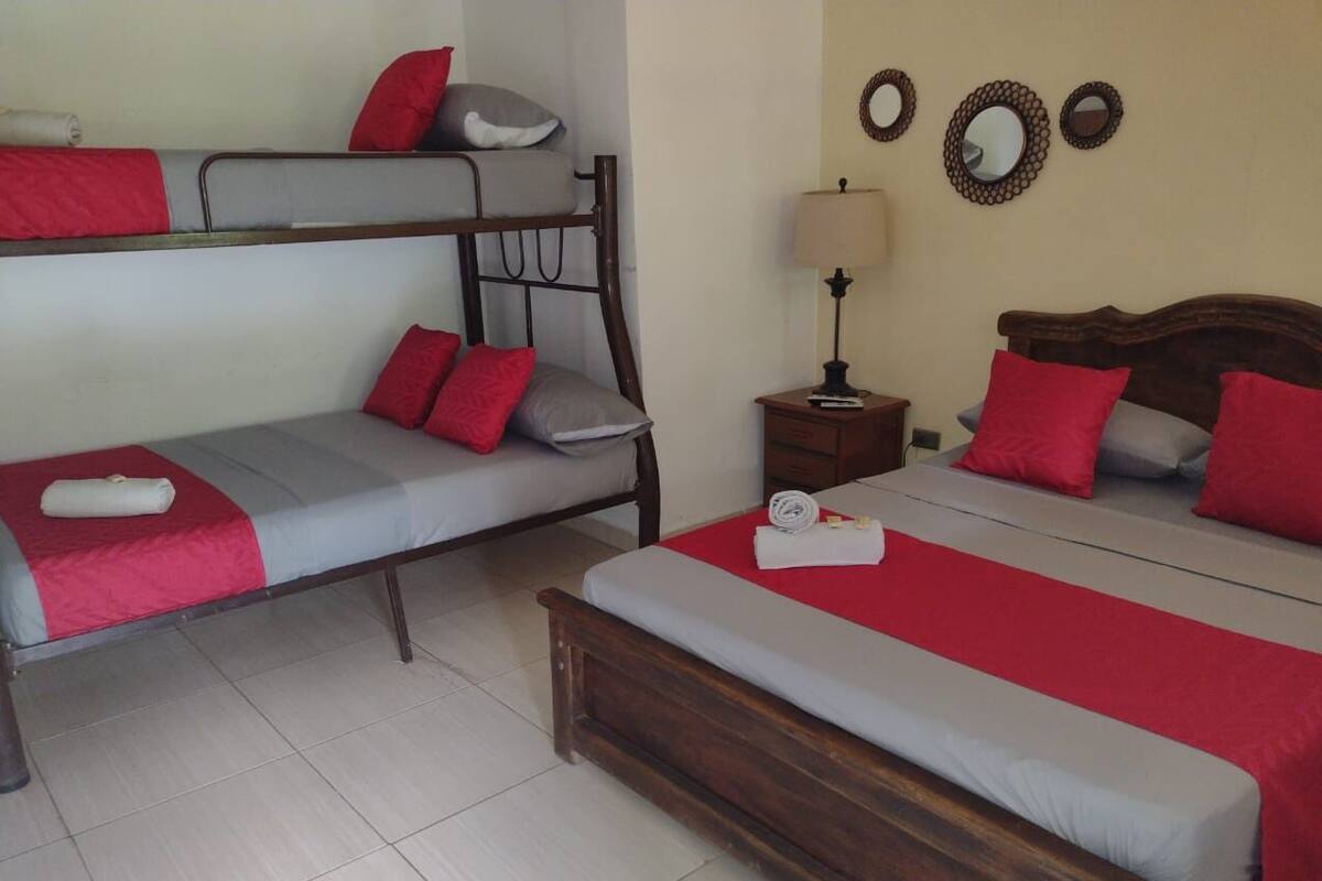 Um quarto com duas beliches, uma mesa de cabeceira com luminária, e uma parede com três espelhos.