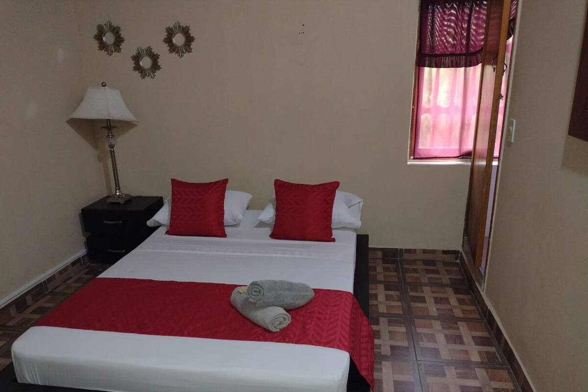 Quarto de hotel com cama, mesa de cabeceira, luminária e janela com cortinas.