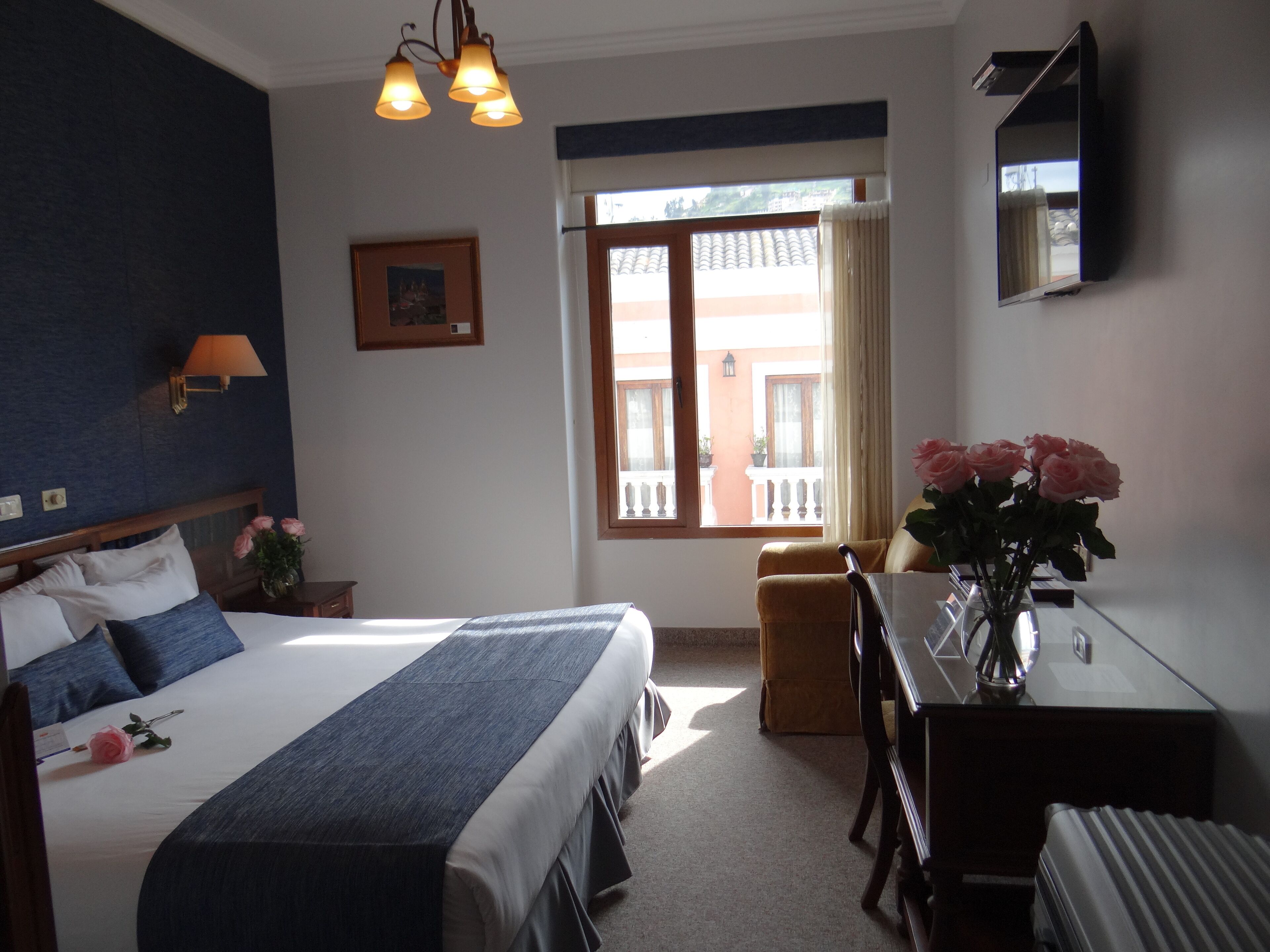 Quarto de hotel com cama, mesa de cabeceira, escrivaninha, cadeira e janela com vista para edifícios.